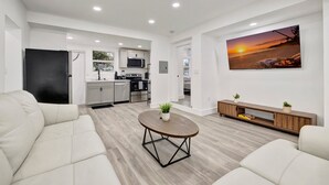 Uma sala de estar moderna com um sofá branco, uma mesa de centro e uma cozinha ao fundo.