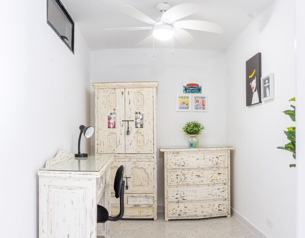 Um quarto com um ventilador de teto branco, um armário branco com portas de vidro, uma mesa pequena com abajur e uma planta sobre uma mesinha lateral.