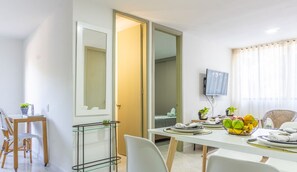 Uma sala de jantar com uma mesa posta para dois, uma TV na parede, e vista para um quarto por uma porta aberta.