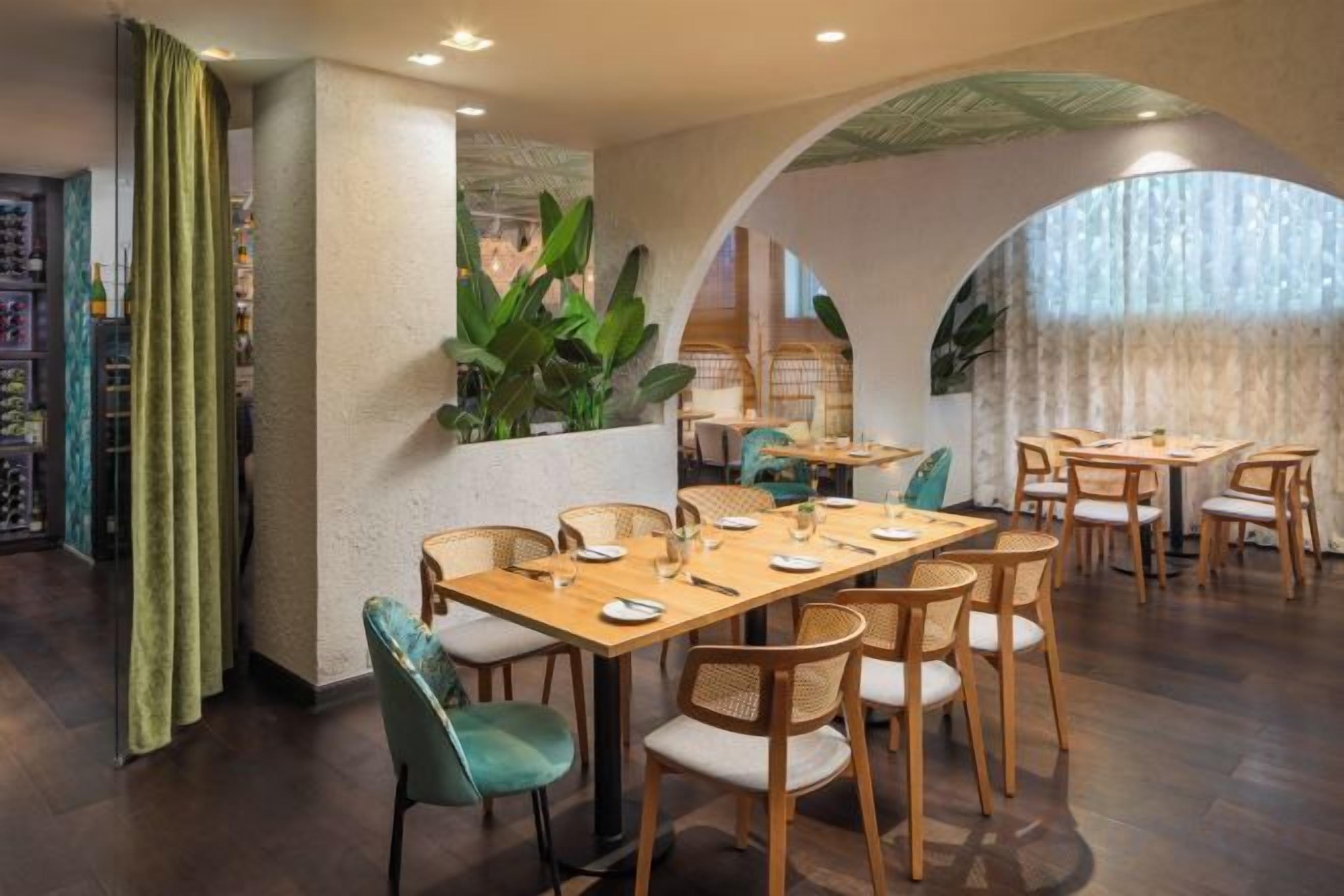 Um restaurante com uma longa mesa de madeira, cadeiras, uma cortina verde e uma planta.