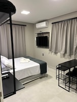 Quarto de hotel pequeno com cama, uma mesinha, televisão fixada na parede e aparelho de ar condicionado.