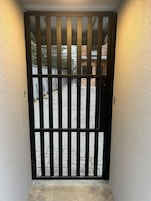 Um portão de metal preto com barras verticais e tela de proteção, dando acesso à entrada de um edifício.