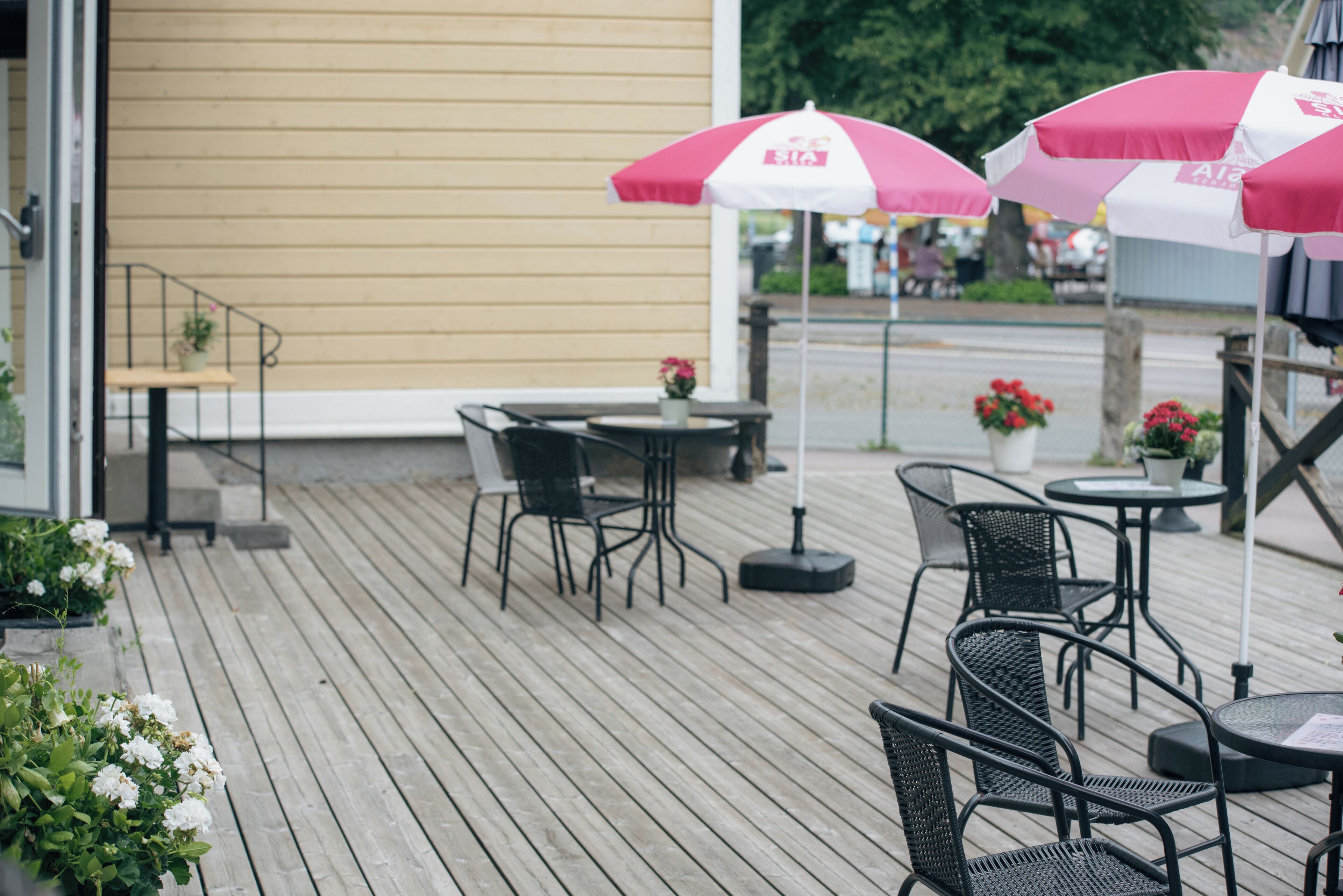 Um terraço ao ar livre com piso de madeira, cadeiras e mesas de metal preto. Há dois guarda-sóis com design rosa e branco.