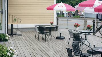 Um terraço ao ar livre com piso de madeira, cadeiras e mesas de metal preto. Há dois guarda-sóis com design rosa e branco.