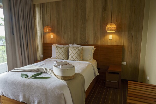 Cama bem arrumada com uma cesta de palha e folhas verdes, cabeceira de madeira, duas luminárias de cabeceira e uma mesa de cabeceira de madeira.