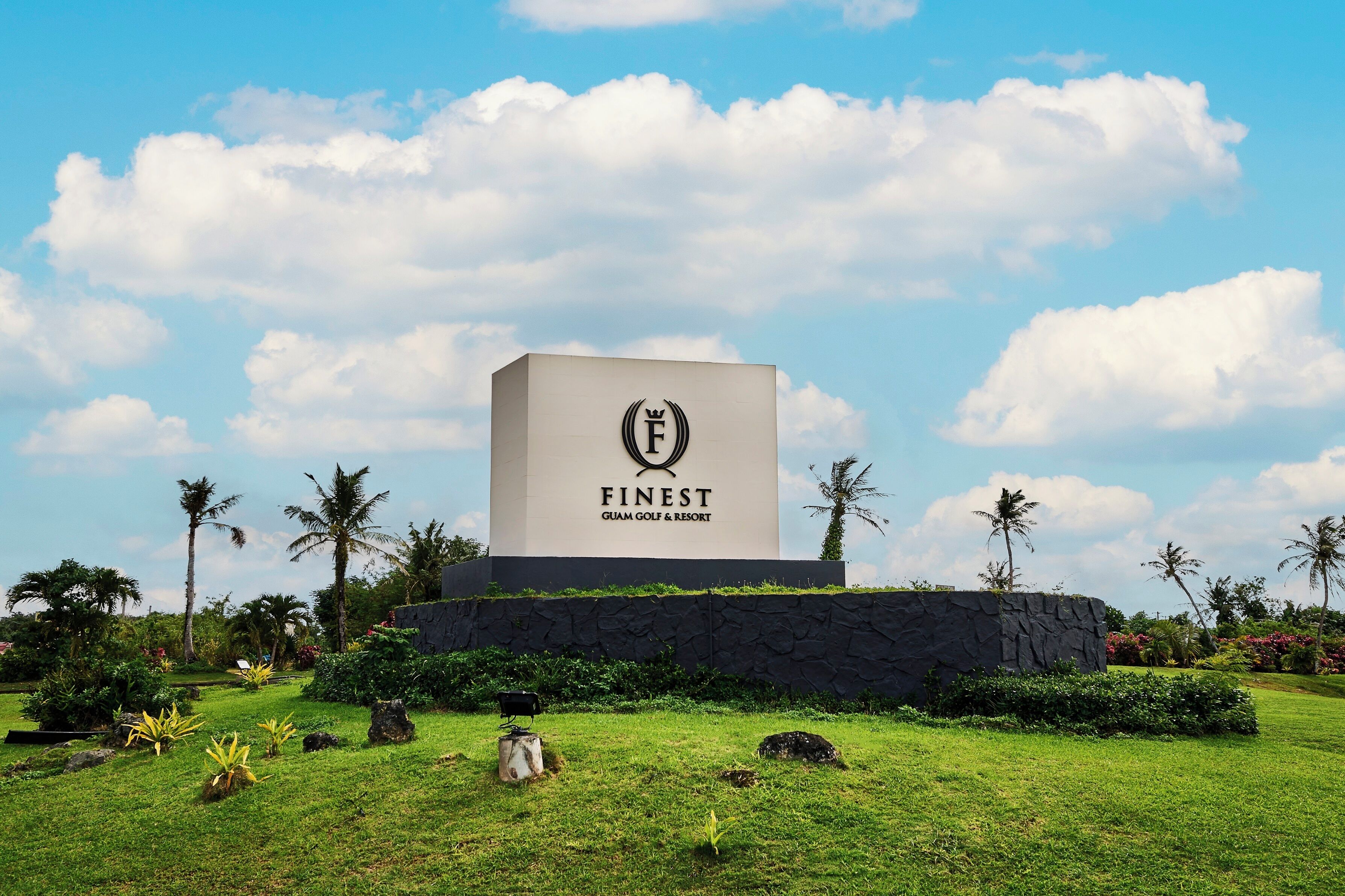 Placa para 'FINEST GUAM GOLF & RESORT', com base de pedra, cercada por vegetação exuberante e palmeiras.