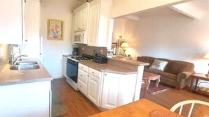 Uma cozinha compacta com pia, fogão e micro-ondas. Há uma área de refeições com mesa e cadeiras, e um sofá na sala de estar.