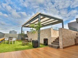 Um terraço no telhado com pérgola, mobiliário de exterior, um contentor de lixo e uma escadaria.