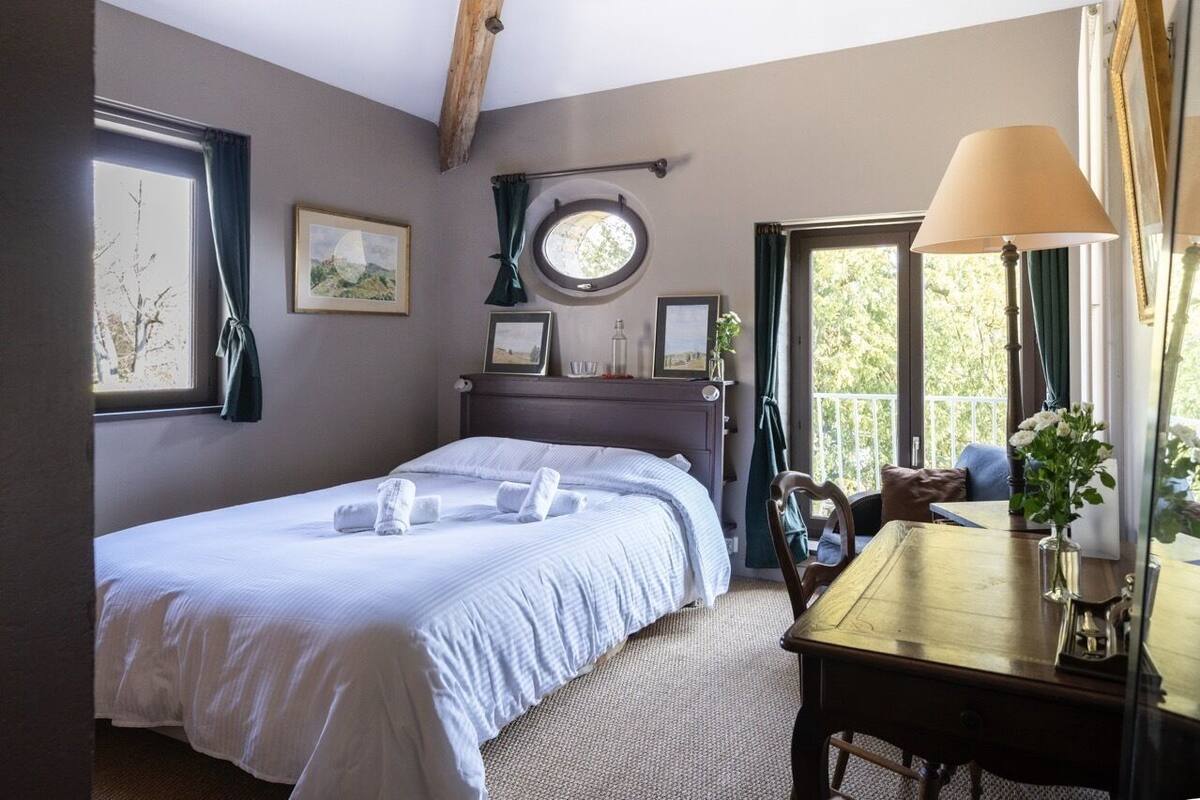 Quarto com uma cama, espelho redondo, mesa com abajur e janela com cortinas.
