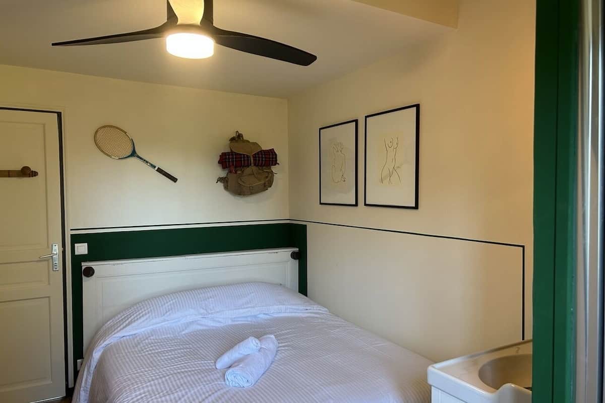 Cama bem arrumada com toalha dobrada, ventilador de teto e raquete de tênis presa na parede.
