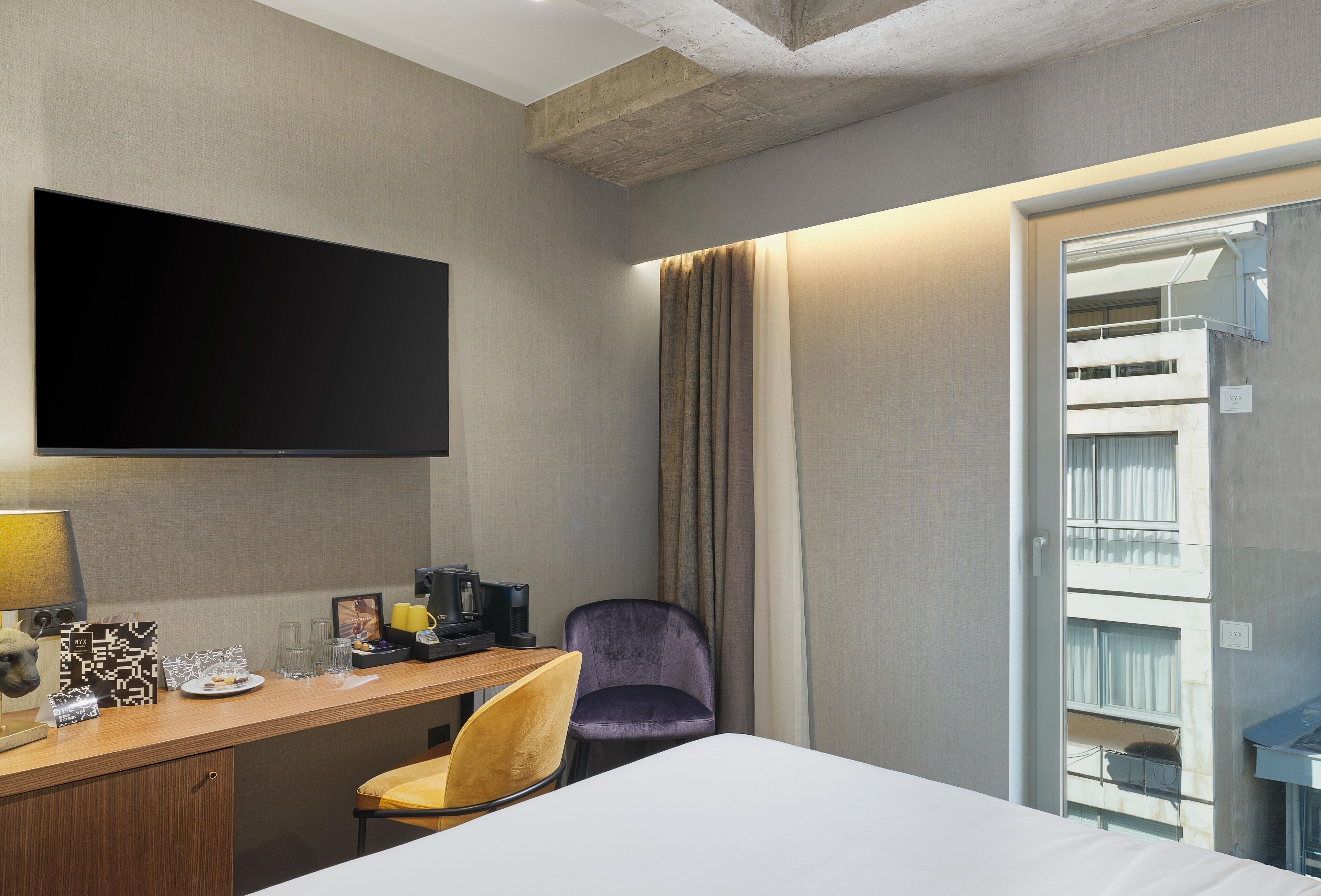 Quarto de hotel com uma cama, uma escrivaninha com televisão de tela plana, uma cadeira e uma janela com cortinas.