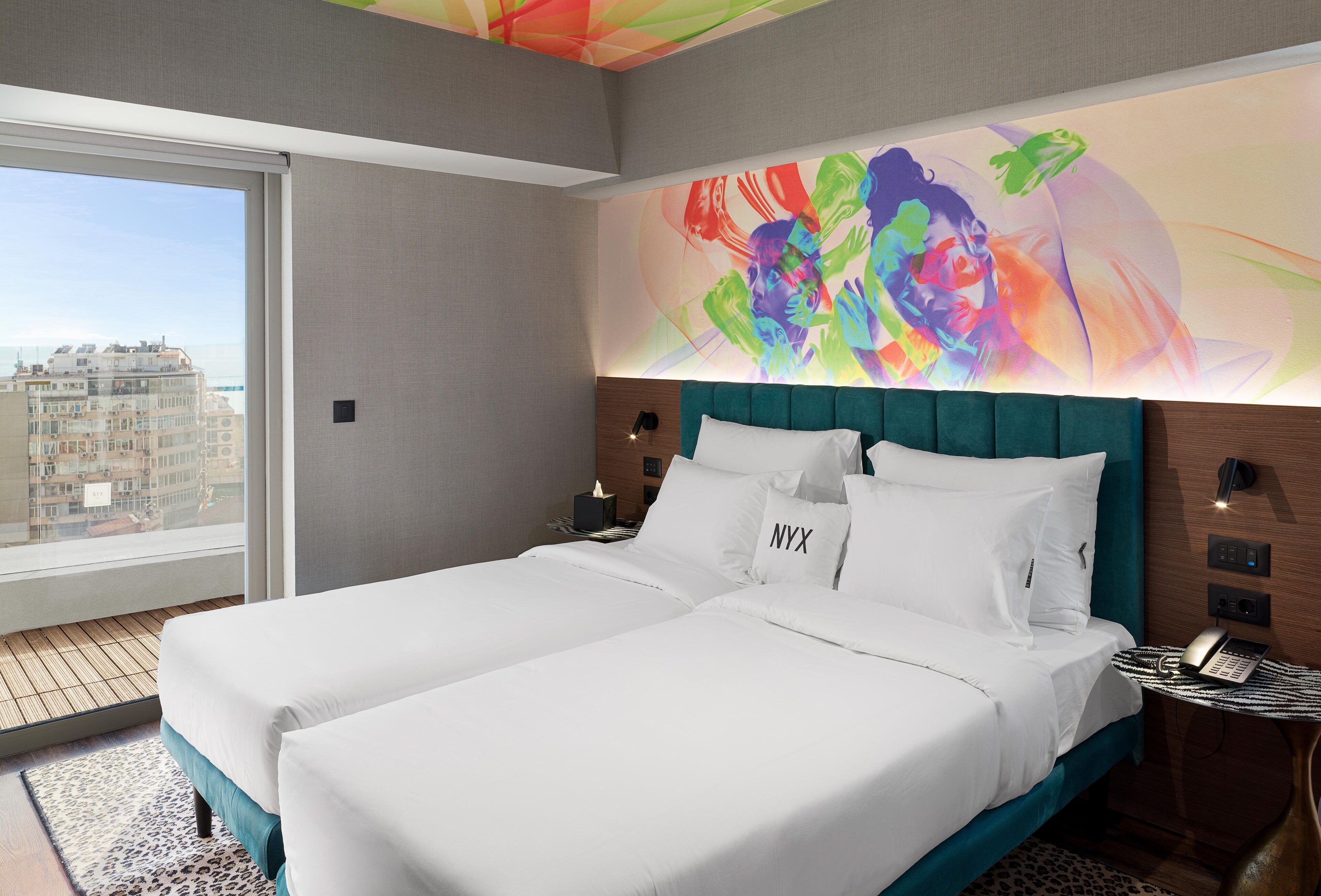 Quarto de hotel com uma cama grande, vista da cidade e um quadro abstrato colorido na parede.
