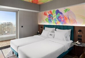 Quarto de hotel com uma cama grande, vista da cidade e um quadro abstrato colorido na parede.