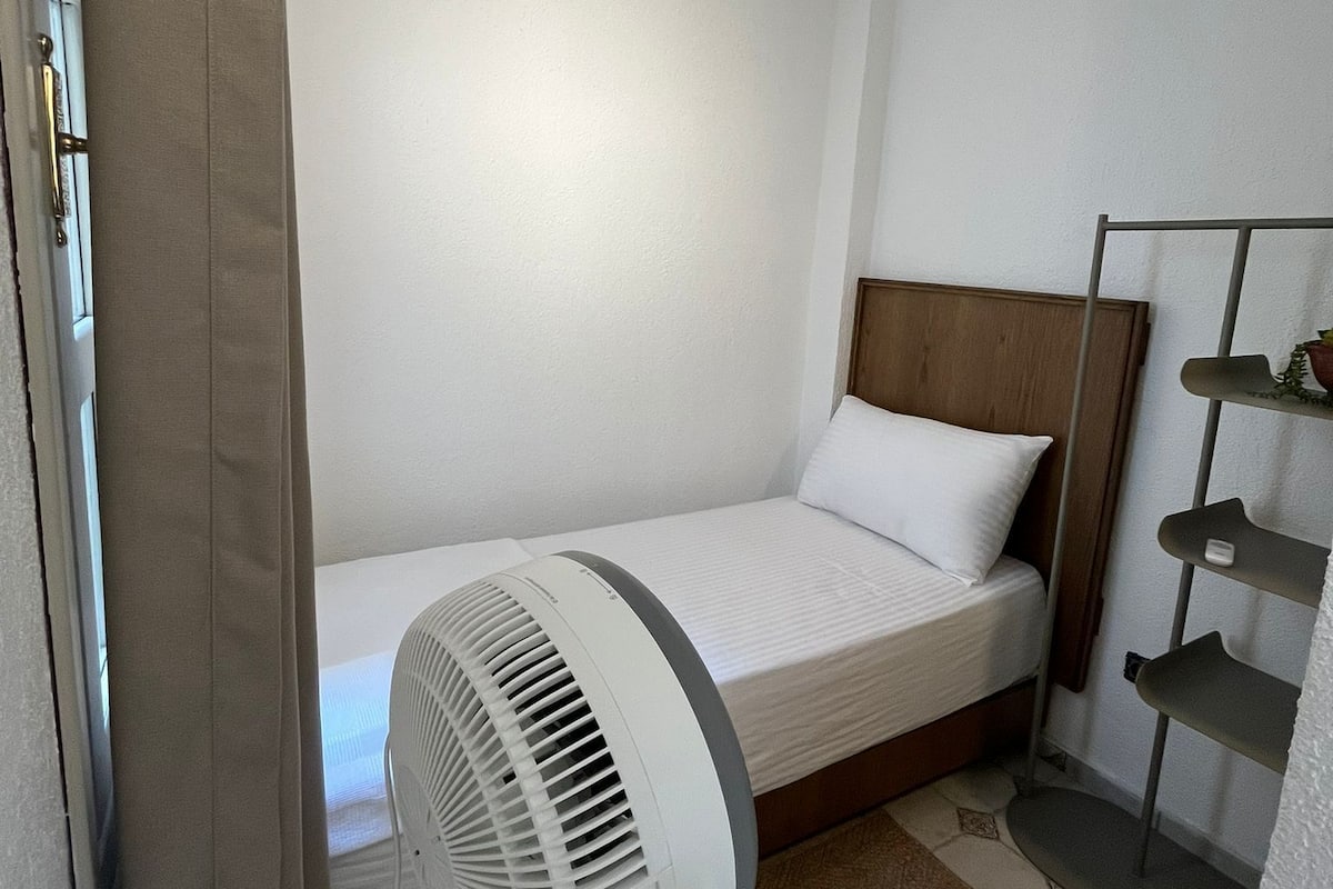 Quarto pequeno com uma cama de solteiro, um ventilador branco, uma cabeceira de madeira e um pequeno armário.