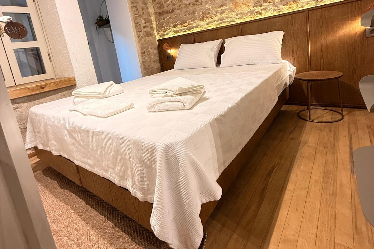 Uma cama bem arrumada com roupa branca, uma mesinha redonda e uma parede de pedra com um aparelho de ar condicionado em cima.