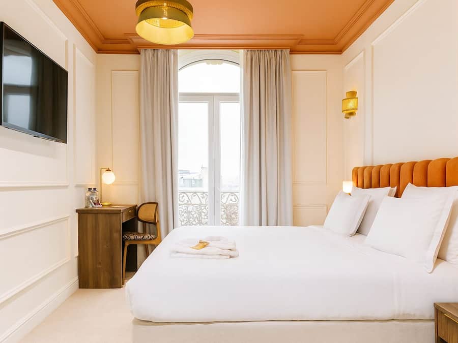 Quarto de hotel com uma cama grande, uma televisão presa na parede, uma escrivaninha com abajur e vista para a cidade pela janela.