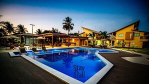 Uma área de piscina com espreguiçadeiras e guarda-sóis, uma placa com a inscrição 'Stay Fall' e um edifício de design moderno.