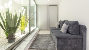 Uma sala de estar moderna com um sofá cinzento, uma porta de vidro que dá para o exterior e uma planta grande.