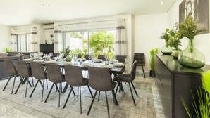 Uma área de refeições moderna com uma mesa grande posta para seis pessoas, rodeada por cadeiras, e uma cozinha ao fundo.
