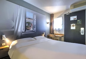 Quarto de hotel com cama, escrivaninha, cadeira, janela com cortinas e porta com calendário e aviso.