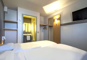 Quarto de hotel com cama, espelho, televisão e banheiro visível por uma porta aberta.