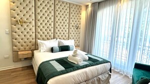 Quarto de hotel moderno com uma cama grande, cabeceira com design acolchoado, mesa de cabeceira e vista para o exterior através de cortinas leves.