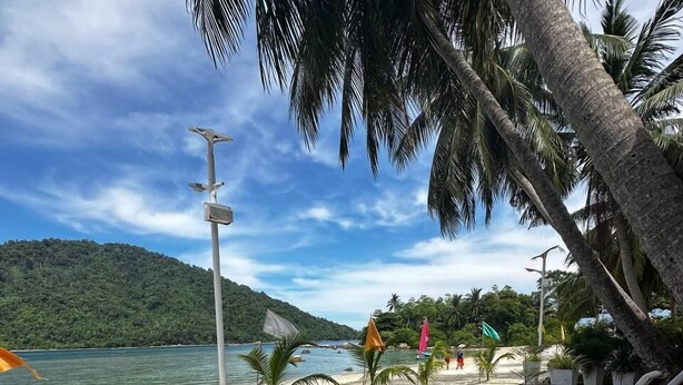 Um deck de madeira com espreguiçadeiras e uma mesa, com vista para uma praia com palmeiras e uma montanha ao fundo.