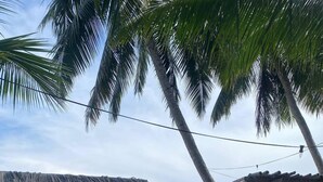 Uma área de estar à beira-mar com teto de palha, deck de madeira e cadeiras de descanso. Uma placa indica o local como 'Verandah Belong Hoom 317'.
