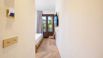 Quarto de hotel com cama, poltrona, escrivaninha, televisão e varanda com vista para palmeiras.