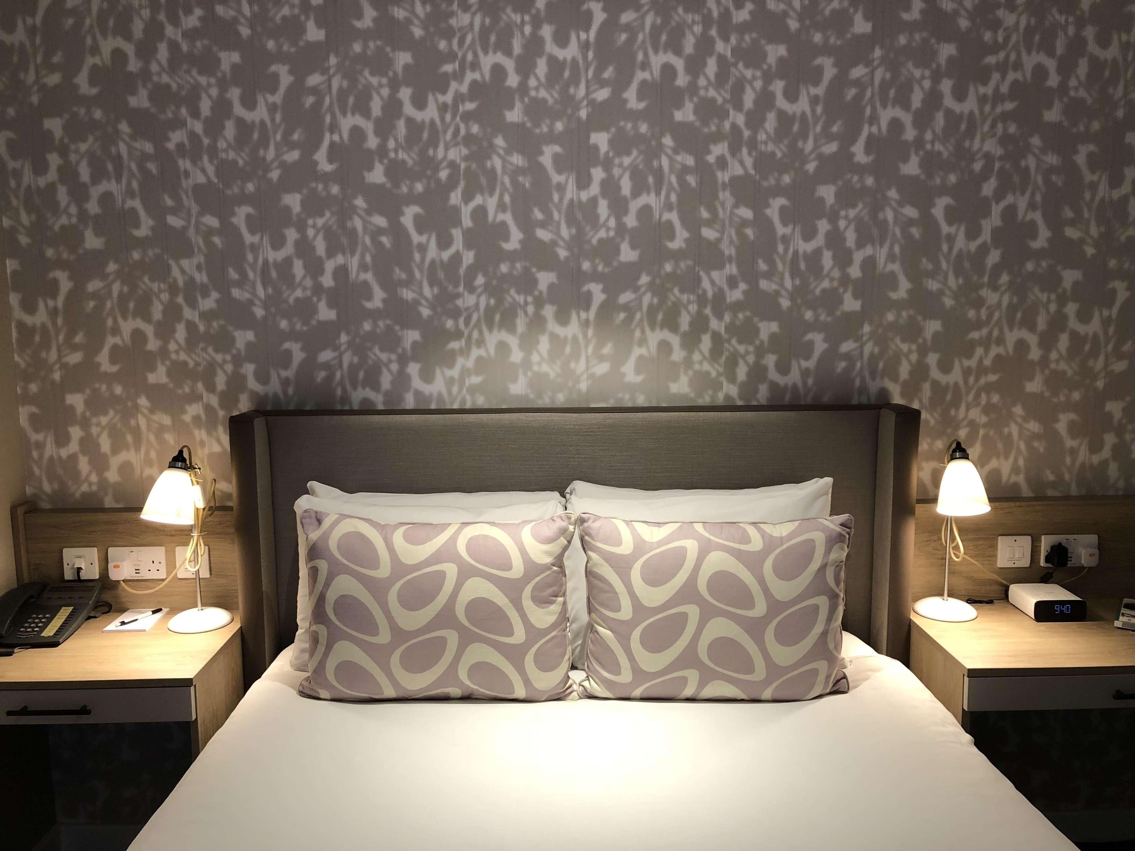 Quarto de hotel com cama, duas luminárias de cabeceira, mesa de cabeceira com telefone, relógio digital e parede estampada.