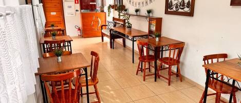 Uma área de jantar com várias mesas e cadeiras de madeira, uma cozinha ao fundo e um banner na parede.