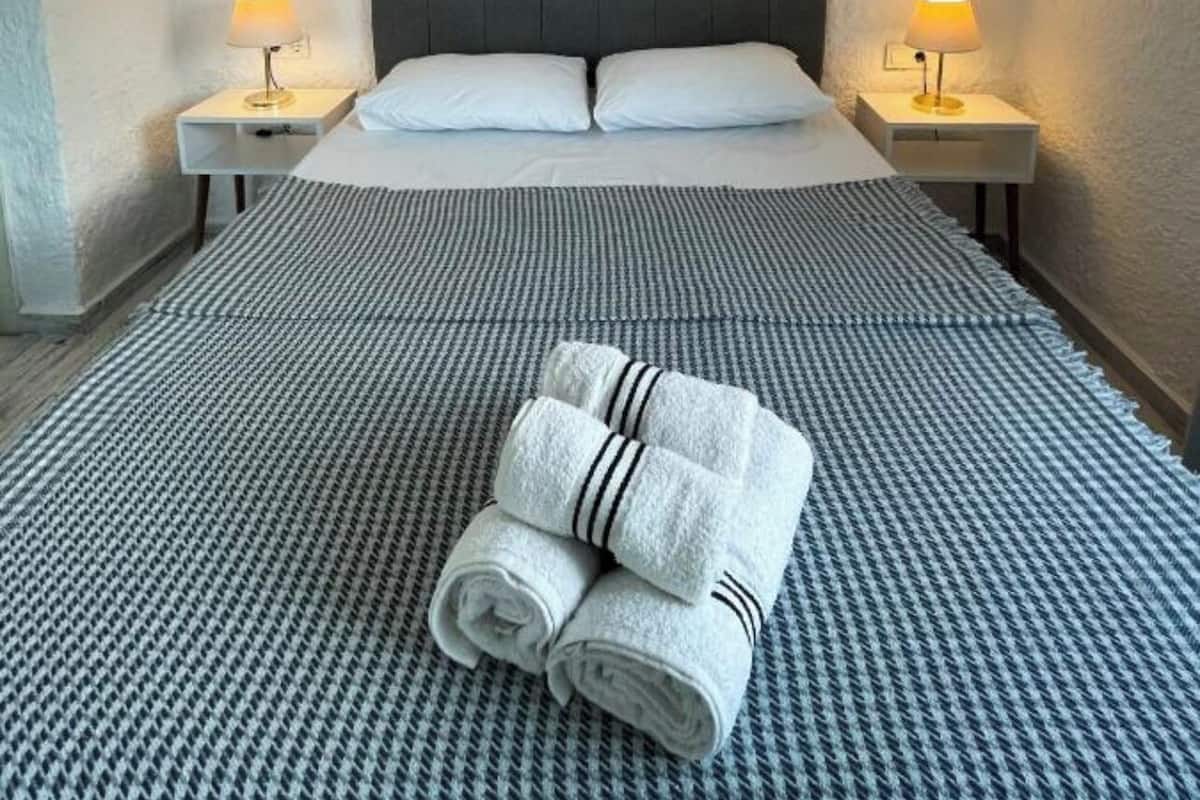 Uma cama bem arrumada com colcha listrada cinza e branca, duas almofadas brancas e um par de toalhas enroladas na cama.