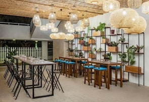 Uma sala de jantar com mesas e cadeiras compridas, luminárias de vime penduradas e prateleiras com plantas em vasos.