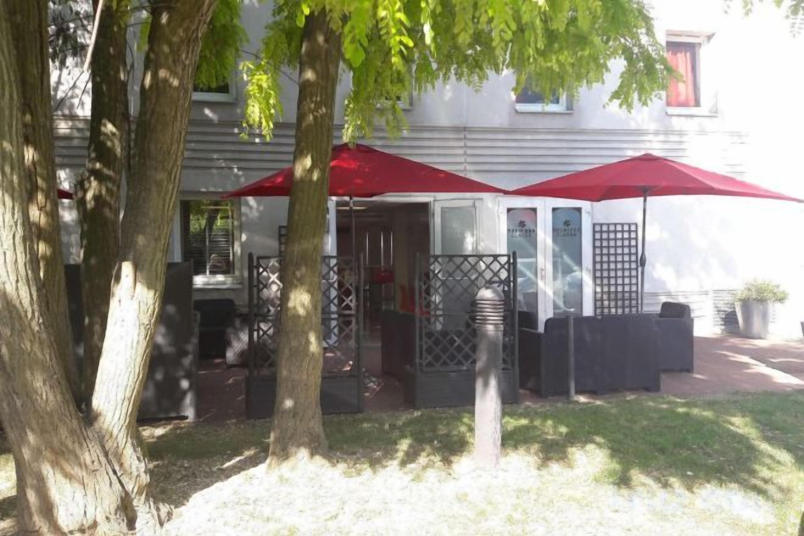 Uma área de pátio com mesas ao ar livre, um guarda-sol vermelho e um edifício ao fundo.