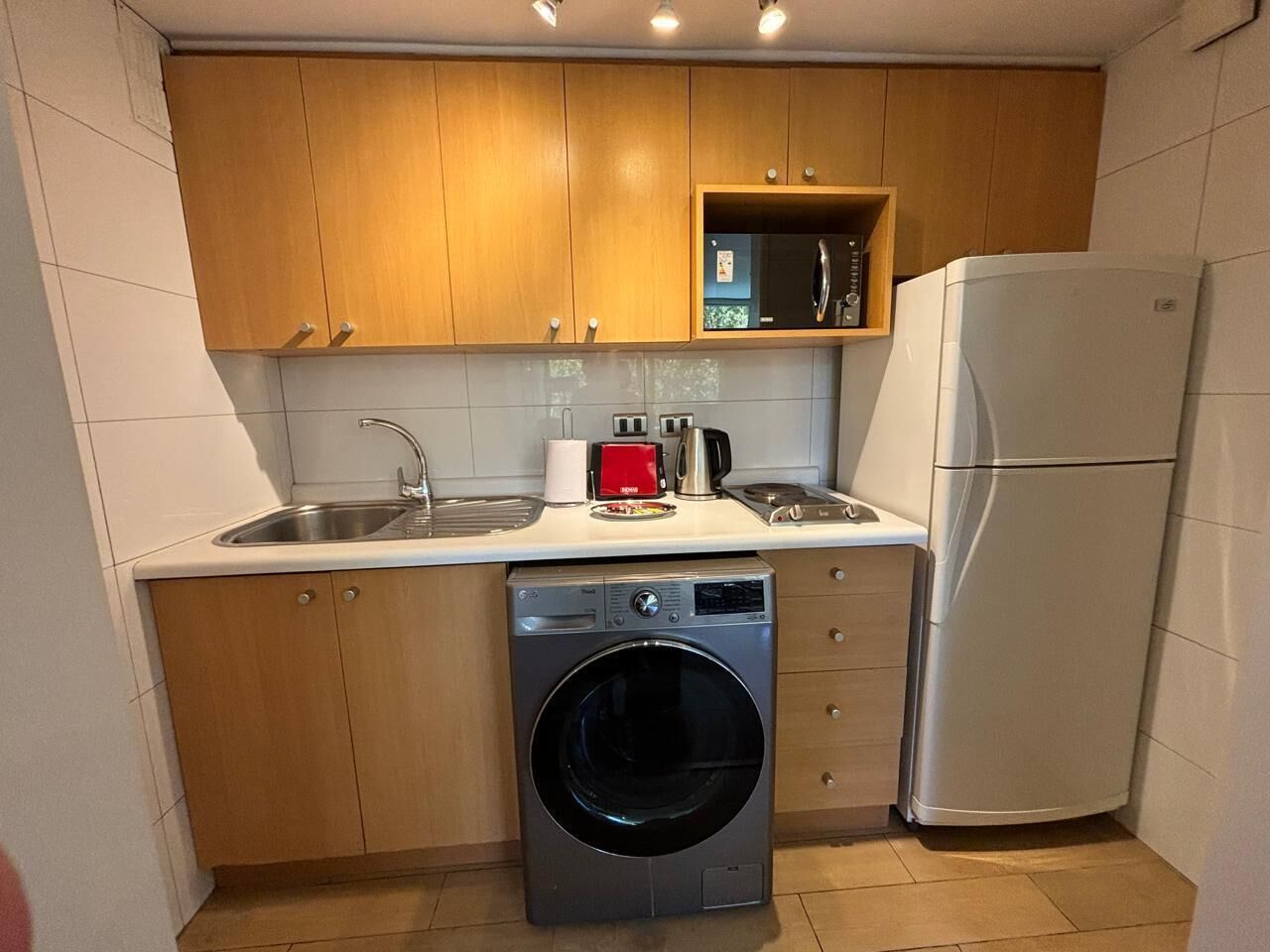 Cozinha compacta com armários de madeira, máquina de lavar, geladeira e micro-ondas.