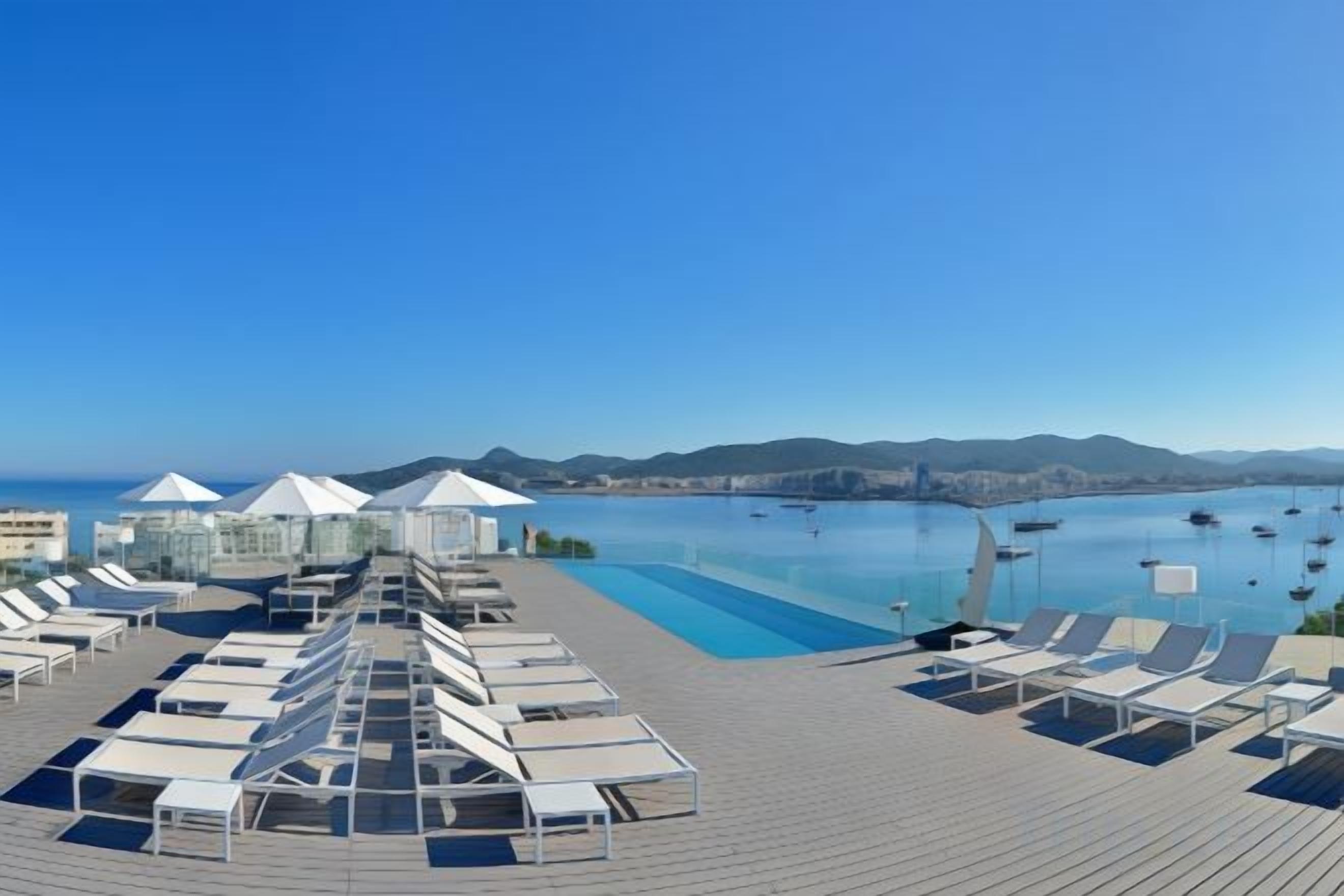 Área de piscina com espreguiçadeiras e guarda-sóis, com vista para um corpo de água com barcos.