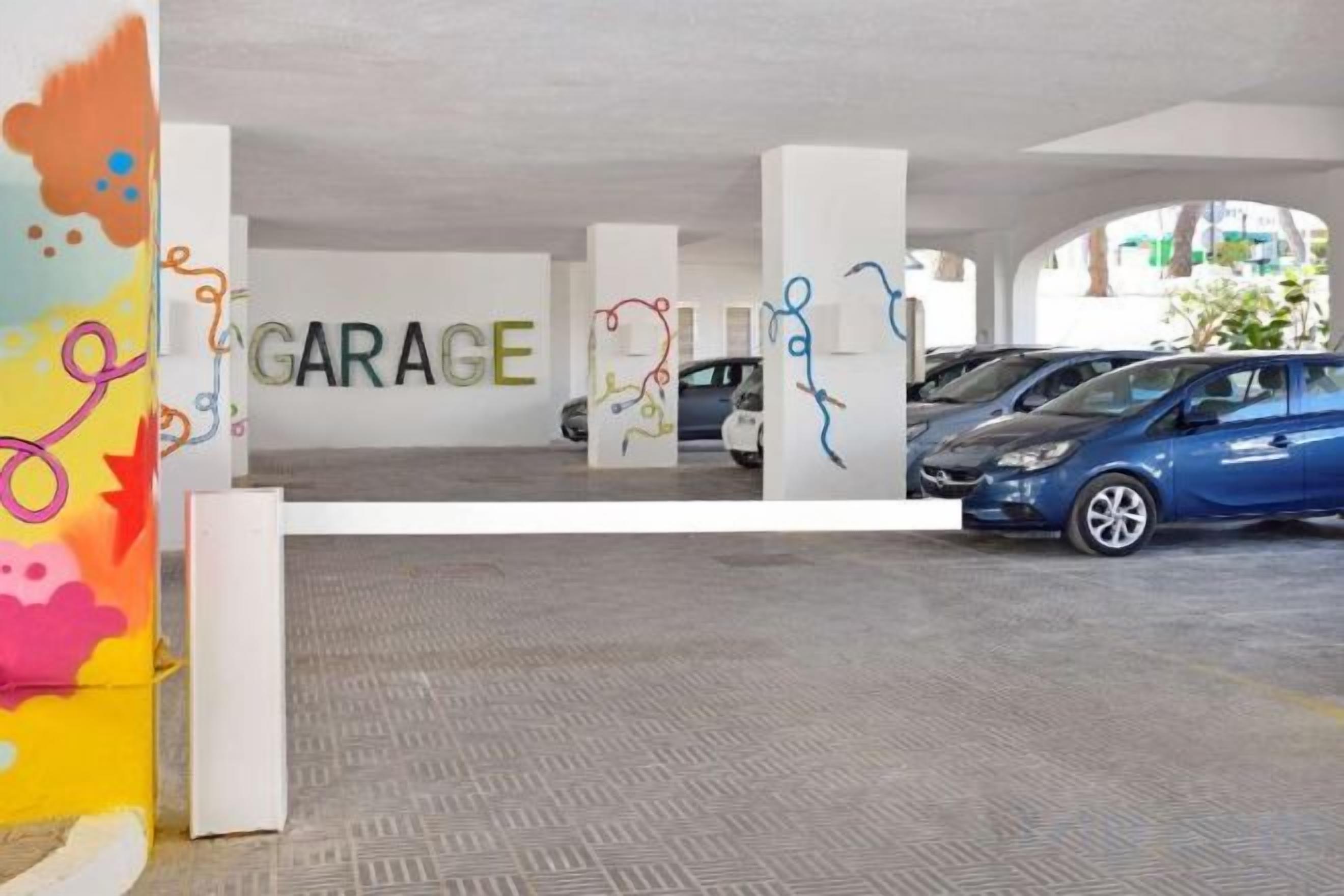 Garagem de estacionamento com vários carros, pilares brancos e uma parede colorida com desenhos abstratos.