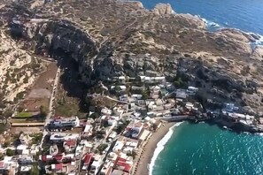 Vista aérea de uma vila costeira com edifícios ao longo da costa, terreno rochoso e um mar azul cristalino.