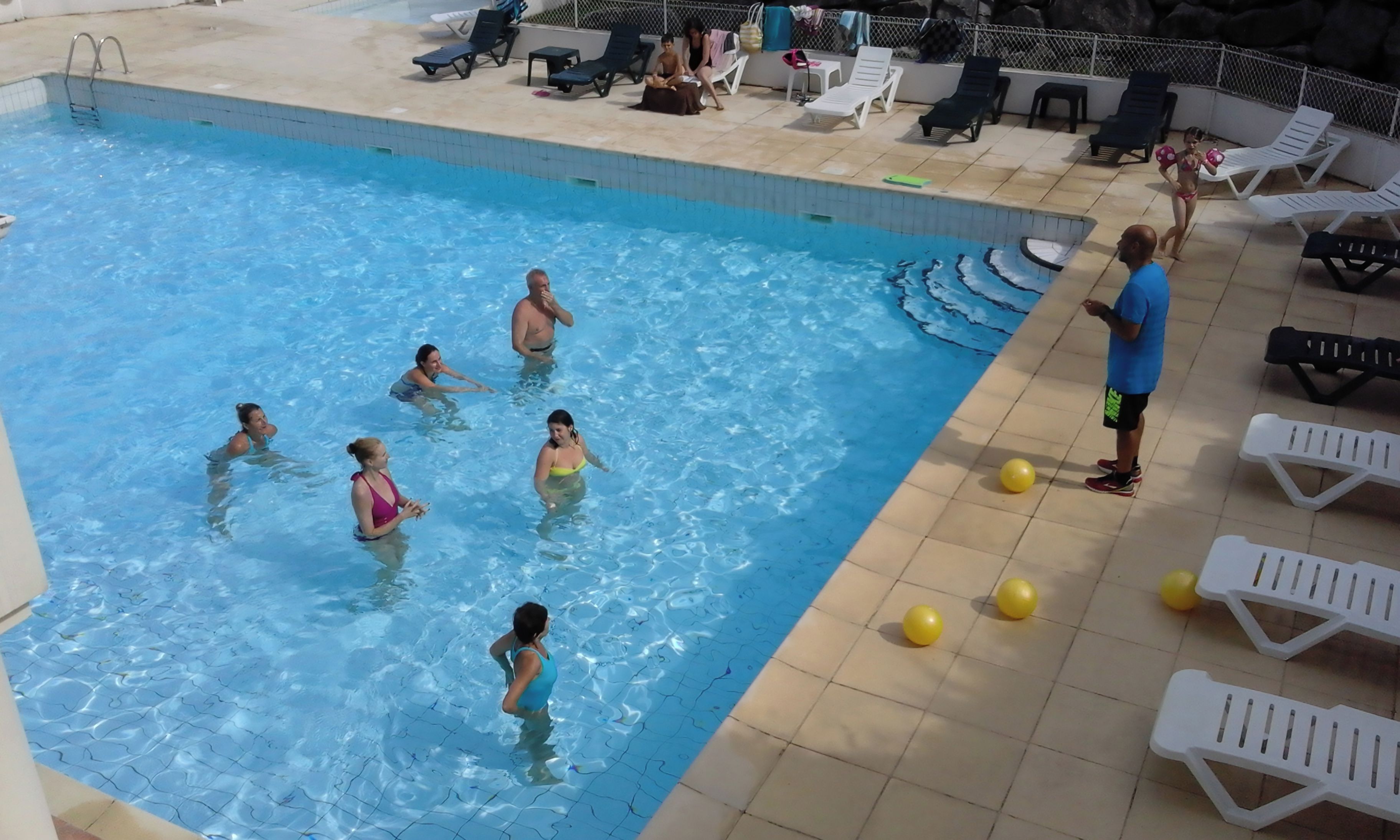 Piscina com pessoas na água, cadeiras de sol e posto de salva-vidas.