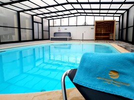 Piscina interna com teto de vidro, um banco de madeira e uma toalha com logotipo.