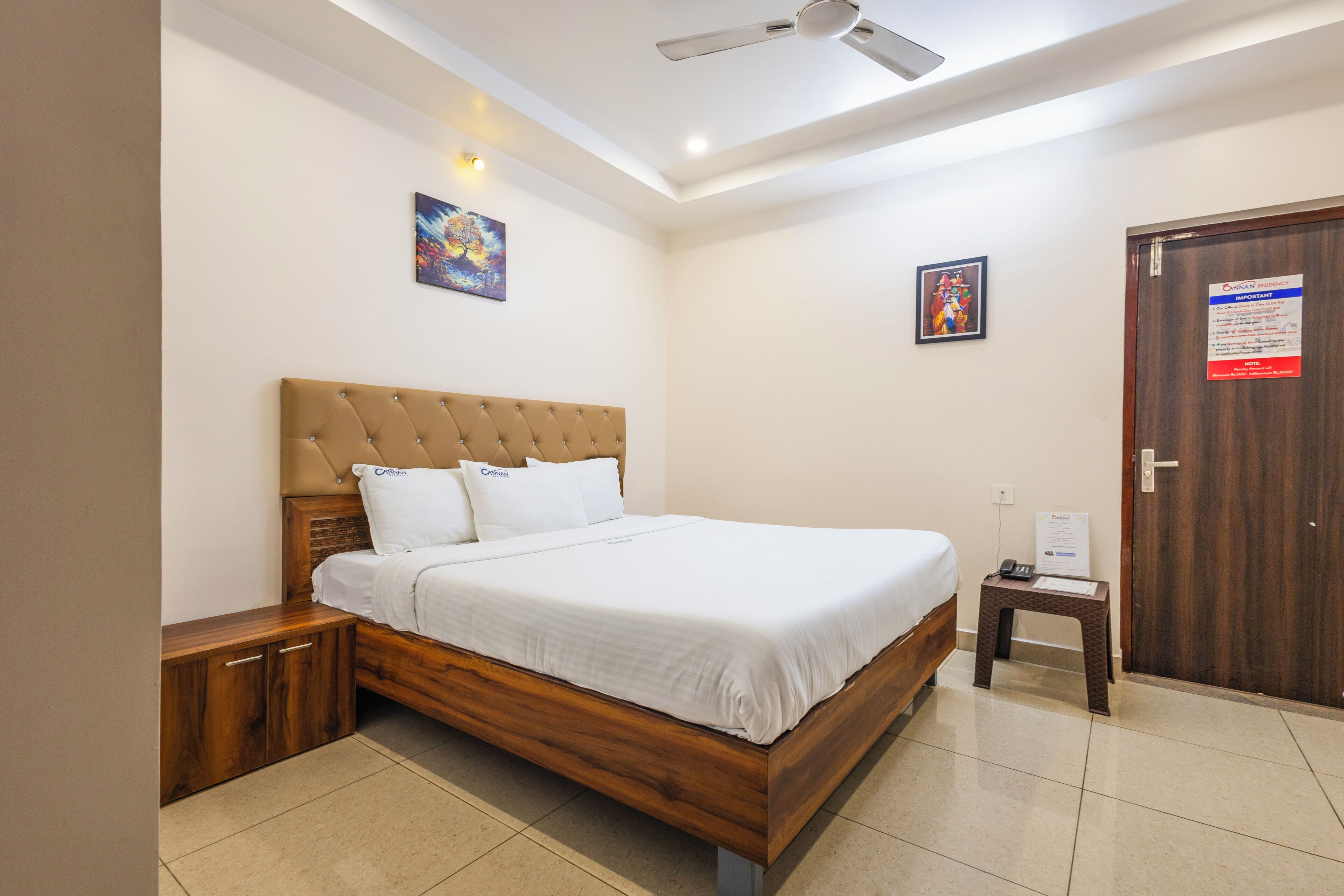 Quarto de hotel com cama de madeira, mesa de cabeceira, ventoinha no teto e porta com um aviso.