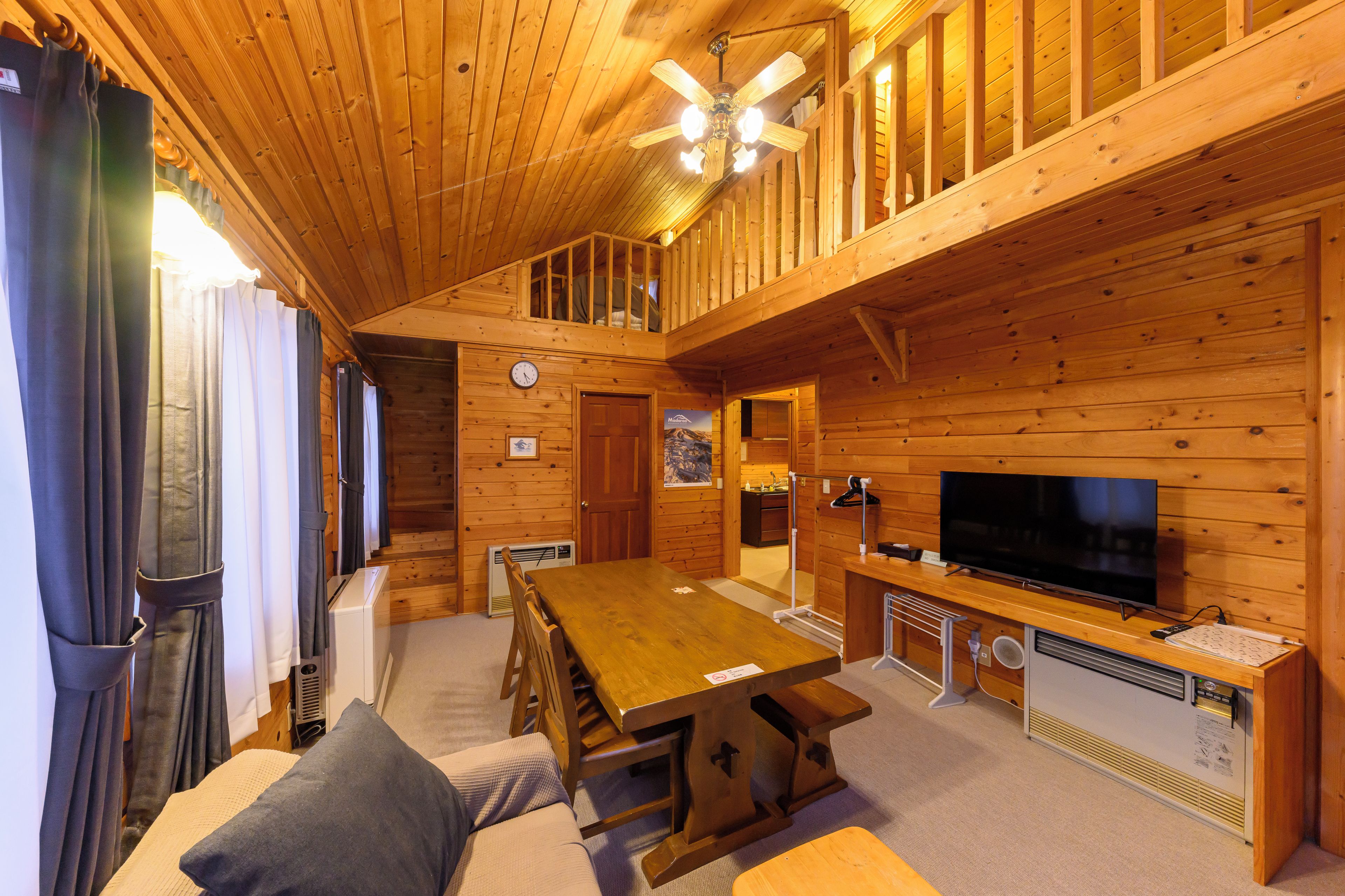 Interior de uma cabana de madeira com mesa de jantar, sofá, televisão e ventilador de teto.