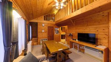 Interior de uma cabana de madeira com mesa de jantar, sofá, televisão e ventilador de teto.