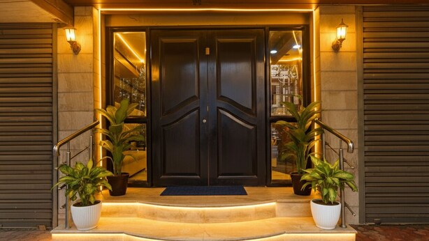 Entrada de hotel com uma porta dupla escura, duas plantas em vasos de cada lado e um tapete azul nos degraus.