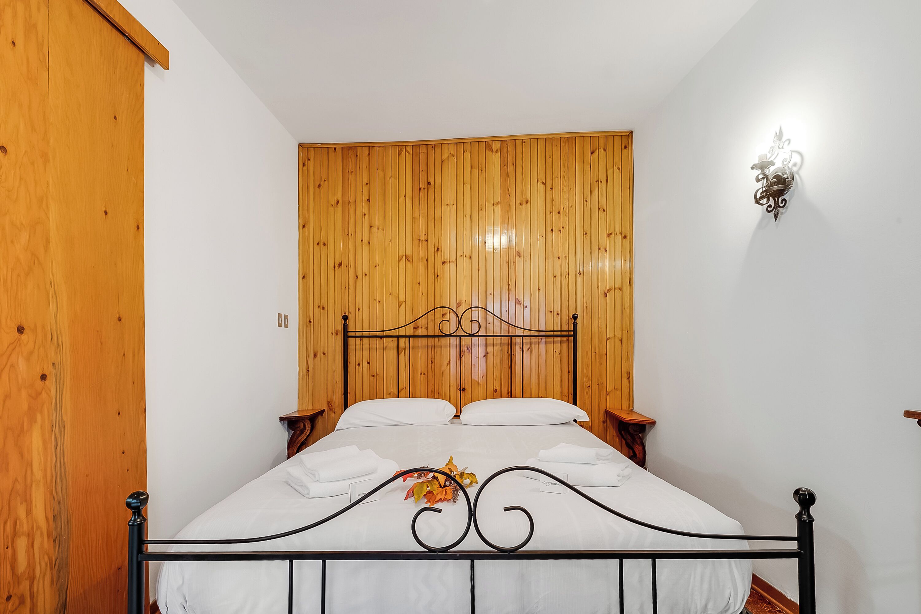 Cama bem arrumada com lençóis brancos e cabeceira decorativa, painéis de madeira na parede e luminária fixada na parede.