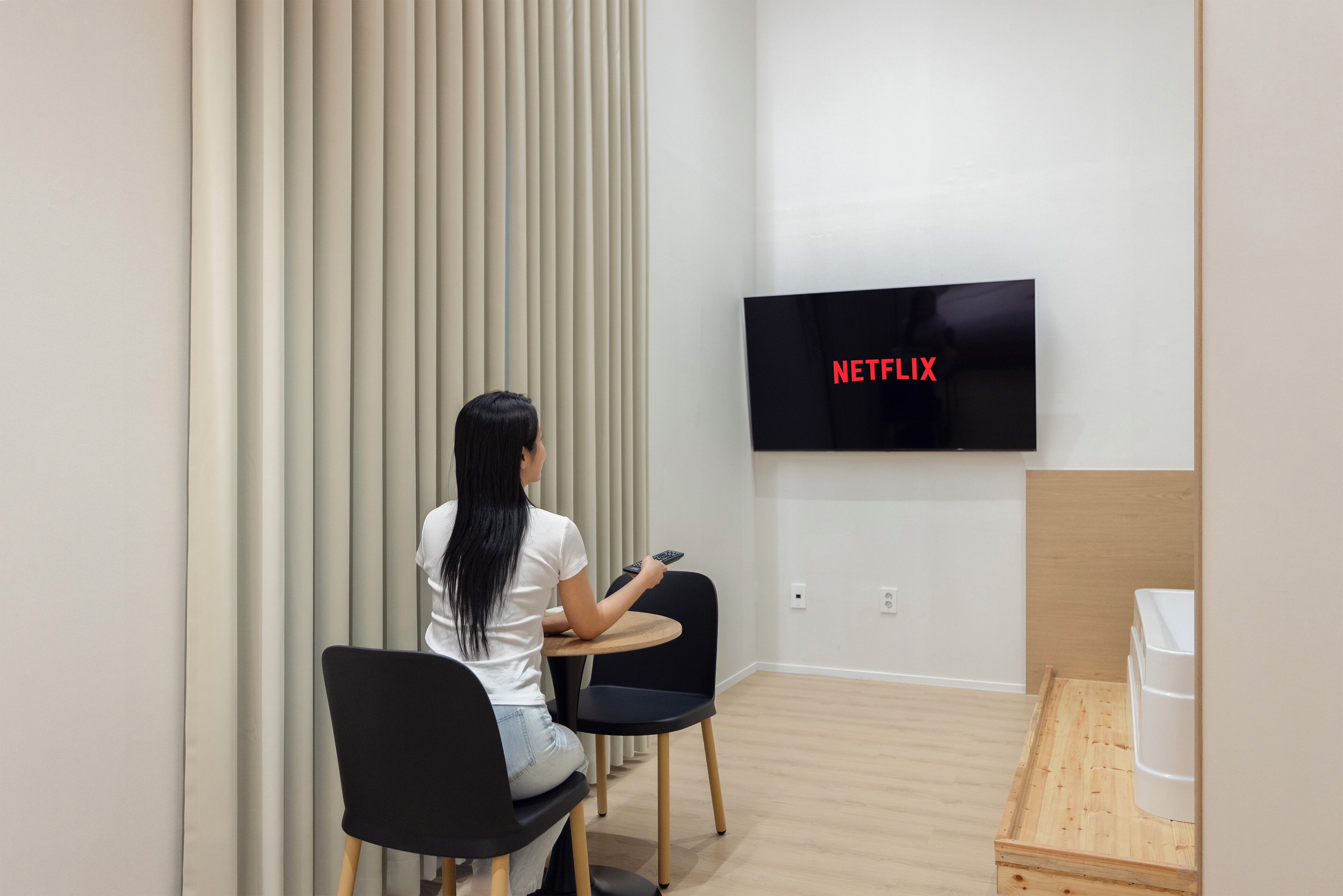 Quarto com televisão exibindo 'NETFLIX', uma mulher sentada à mesa e um armário de madeira.