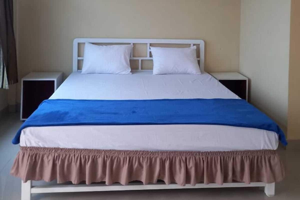 Uma cama de solteiro com cabeceira branca, duas almofadas brancas, colcha azul e saia para a cama marrom. Há duas mesinhas de cabeceira, uma de cada lado da cama.