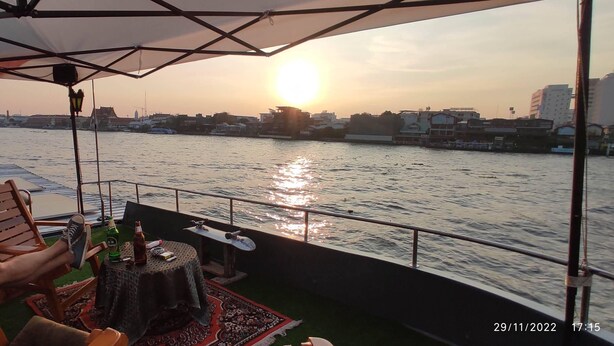 Convés de barco com mesa, cadeiras e vista para o pôr do sol sobre um rio.