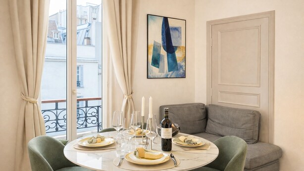Uma área de refeições com uma mesa redonda posta para quatro pessoas, um sofá, um quadro e vista para edifícios através de uma janela.