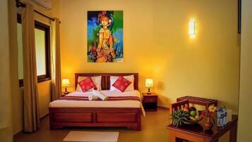 Quarto de hotel com uma cama grande, duas mesas de cabeceira com luminárias, um quadro na parede e uma pequena mesa com frutas e um vaso.